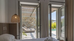 Quarto com uma cama grande, luminária pendente e vista para uma área externa com um muro de pedra e uma piscina.