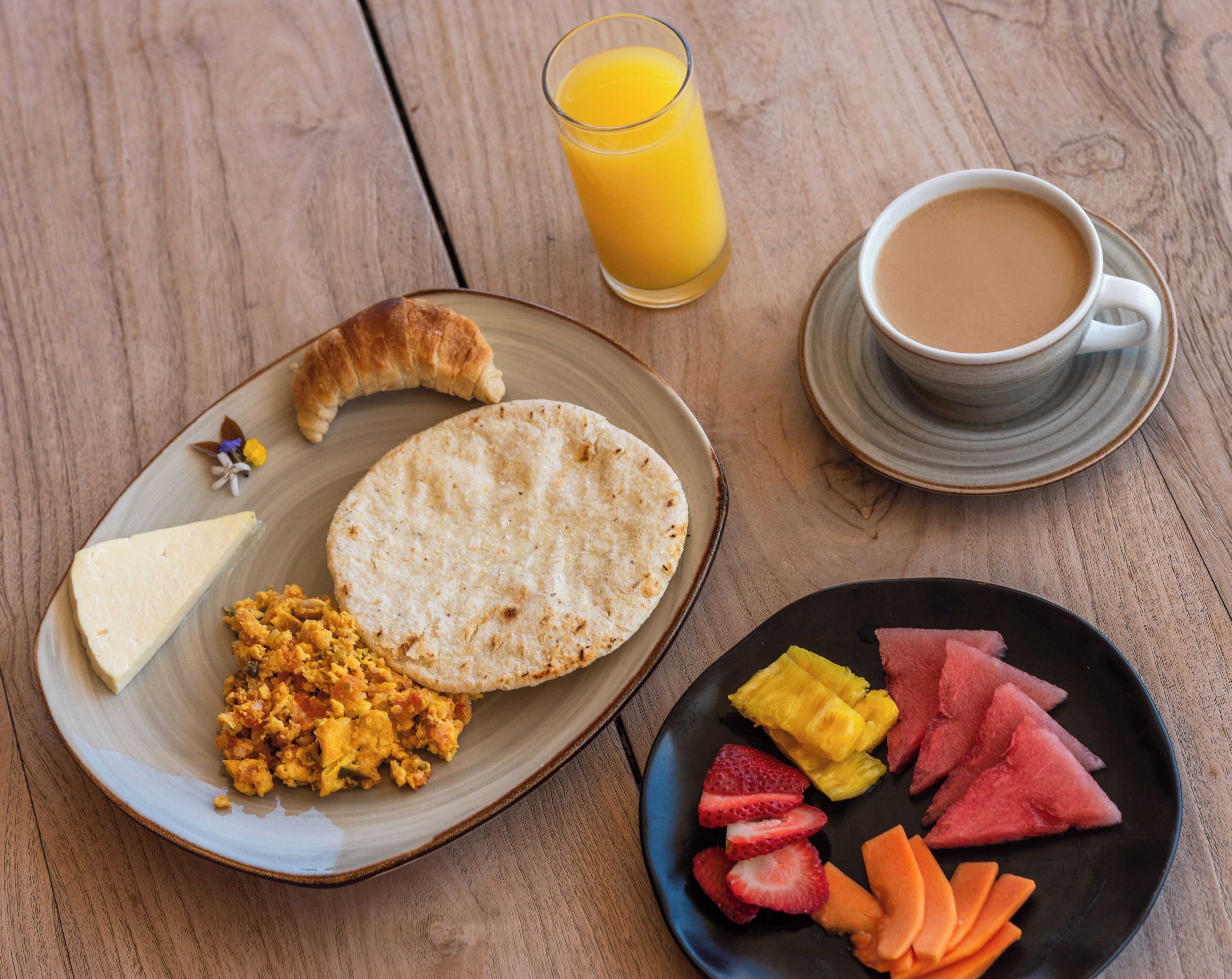 Prato de café da manhã com ovos mexidos, queijo, um croissant e um pão achatado, acompanhado de uma cesta de frutas e uma xícara de café.