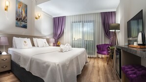 Quarto de hotel com uma cama grande, uma escrivaninha com computador, uma cadeira e vista para o exterior através de cortinas translúcidas.