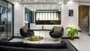 Uma área de recepção moderna com balcão de mármore, duas poltronas pretas com almofadas estampadas, uma mesa de centro de vidro com objetos decorativos e uma planta em vaso.