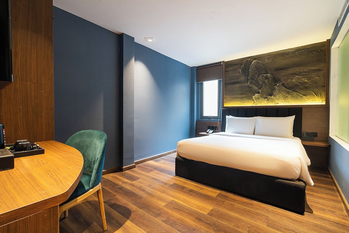 Quarto de hotel com cama, escrivaninha, cadeira e um quadro na parede.