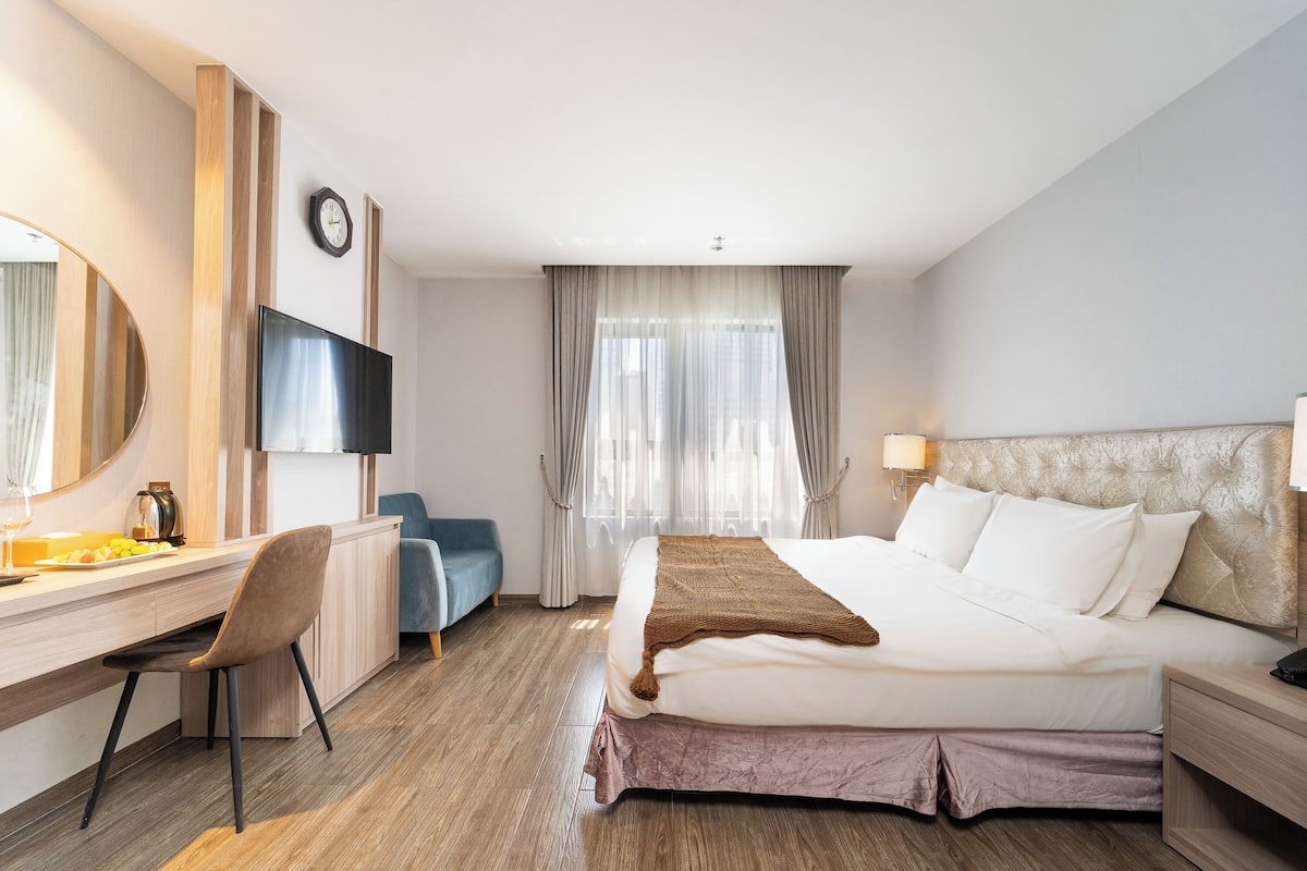 Quarto de hotel com cama, secretária com cadeira, televisão, relógio e janela com cortinas.