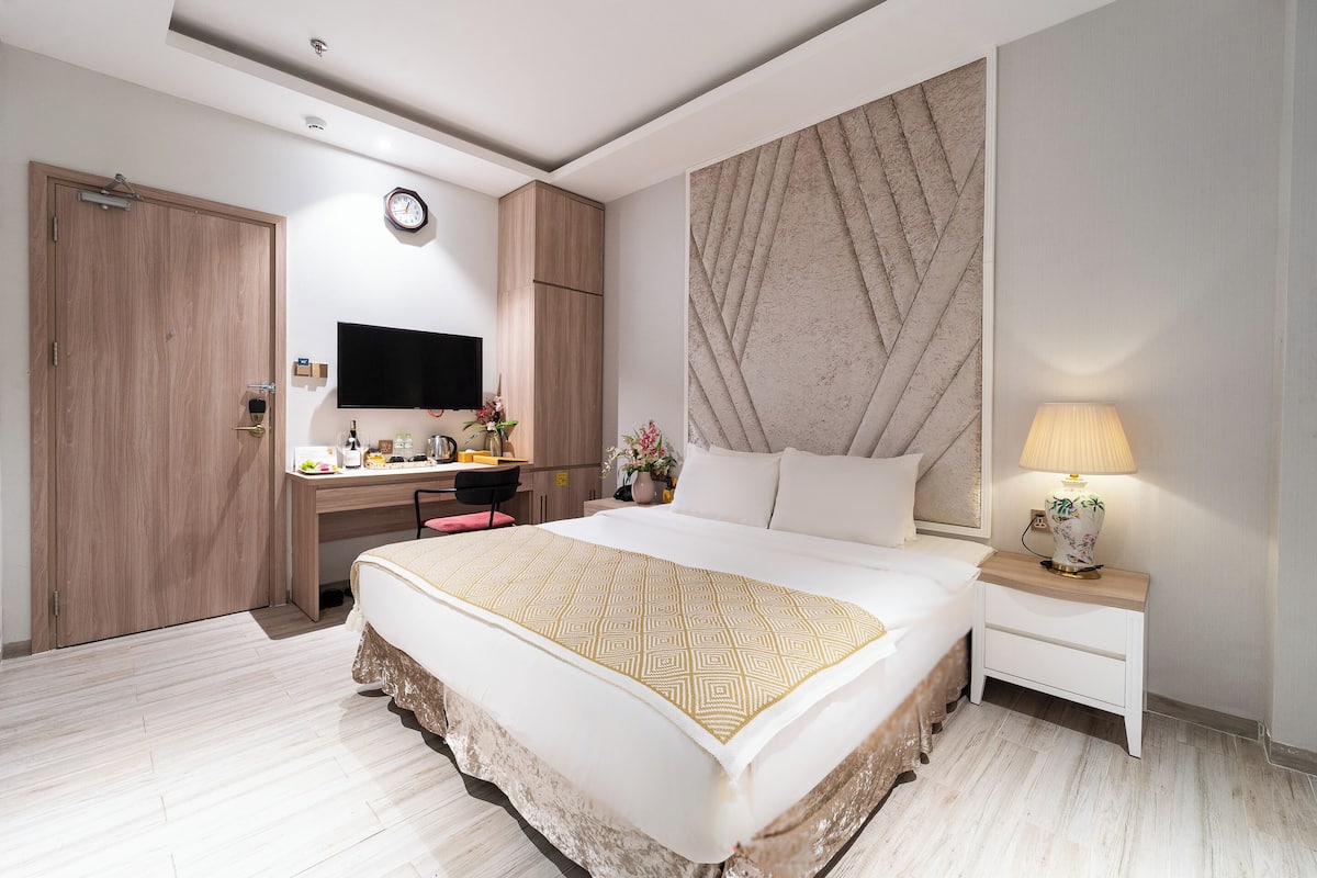 Quarto de hotel com uma cama grande, uma escrivaninha com cadeira, televisão, relógio e mesa de cabeceira com abajur.