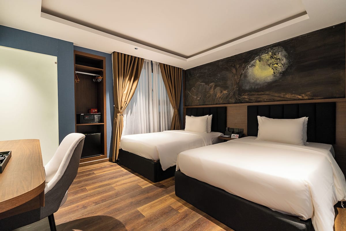 Quarto de hotel com duas camas, uma escrivaninha, uma cadeira, um guarda-roupa e um quadro na parede.