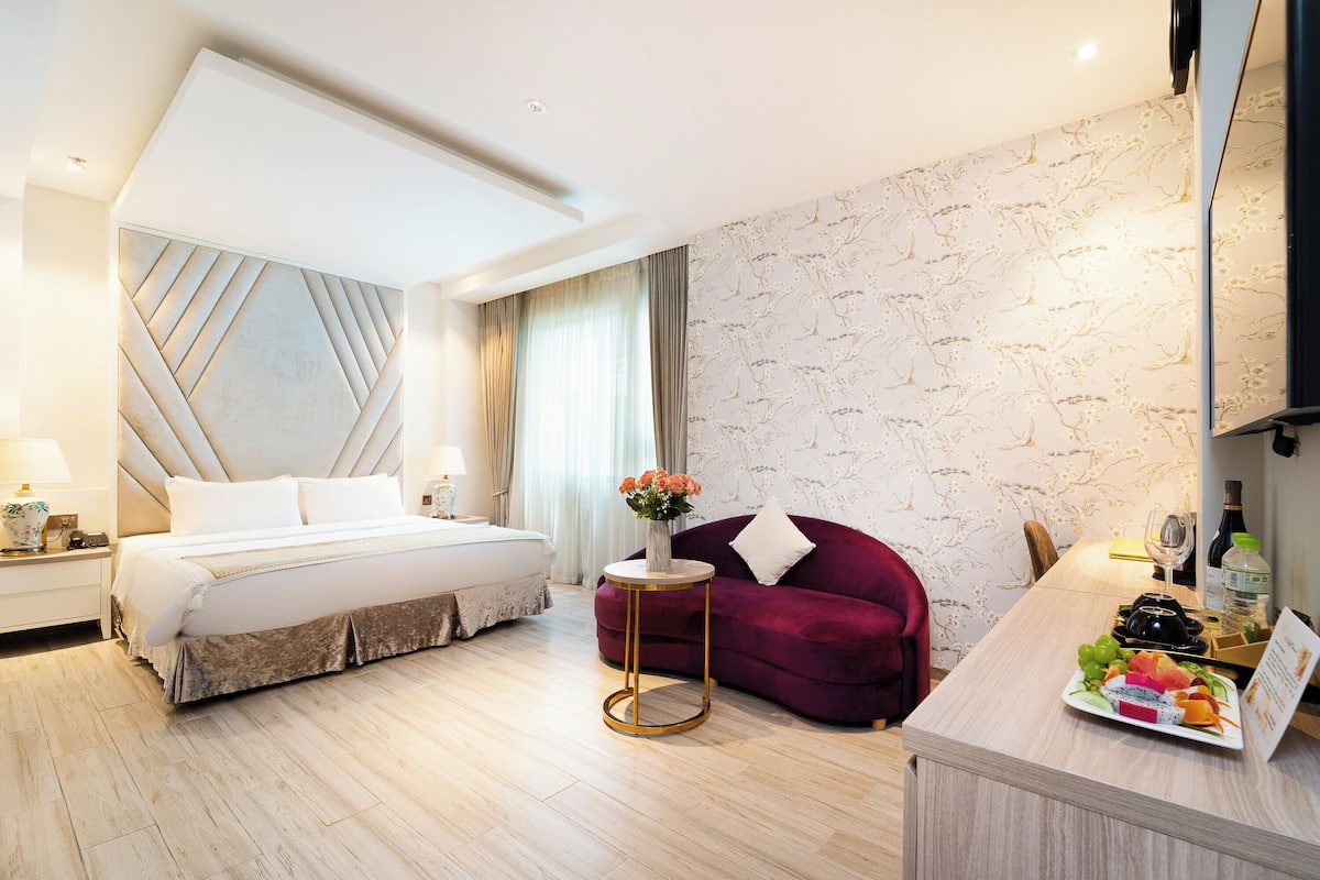 Quarto de hotel moderno com uma cama grande, um sofá vermelho, uma mesinha redonda com um vaso de flores e uma bandeja de café da manhã em cima do aparador.