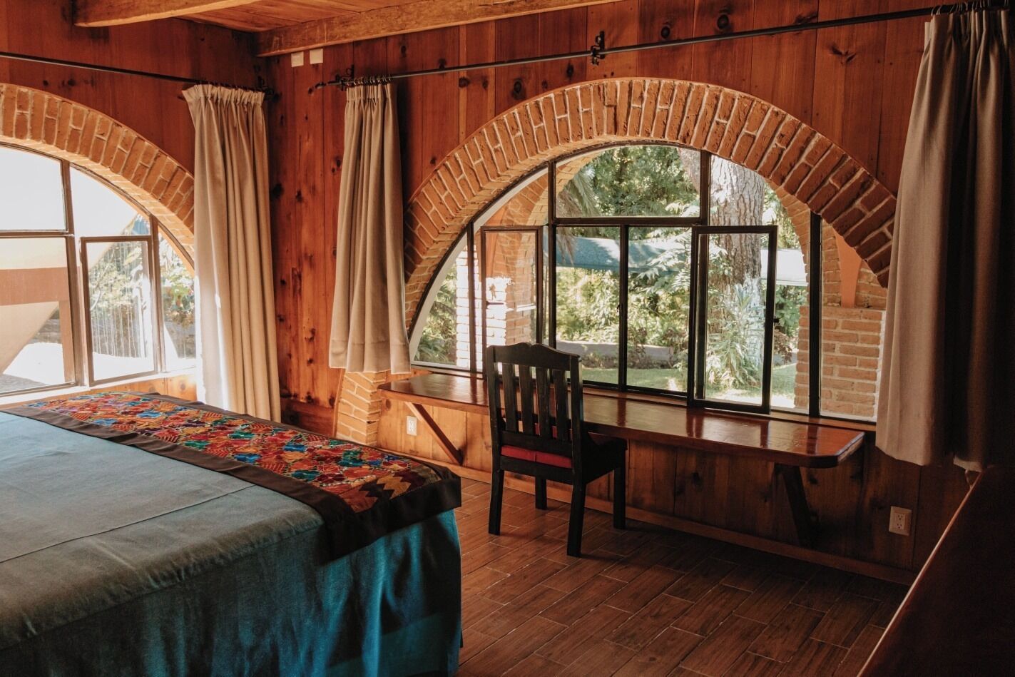 Quarto com cama, uma cadeira e amplas janelas em arco com cortinas.