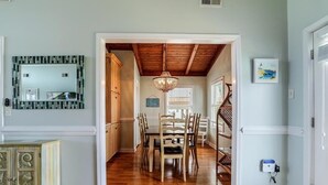 Uma sala de jantar com mesa e cadeiras de madeira, um candelabro e vista para a cozinha através de uma porta aberta.