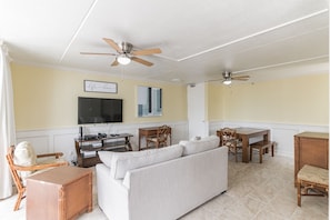 Sala de estar com sofá, cadeiras, mesa de jantar, ventiladores no teto, televisão de tela plana e um quadro na parede.