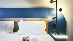 Cama arrumada com lençóis brancos, bandeja com doces e um jarro de café, e luminária fixada na parede.