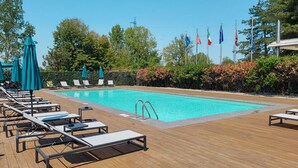 Uma área à beira da piscina com espreguiçadeiras e guarda-sóis, piscina e bandeiras ao fundo.