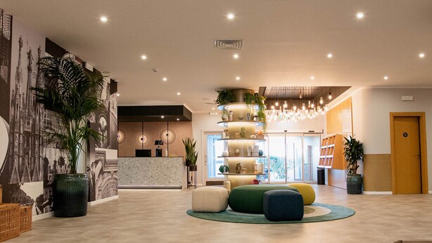 Um hall amplo com balcão de recepção, um arranjo de plantas em níveis e uma área de estar com almofadas coloridas.