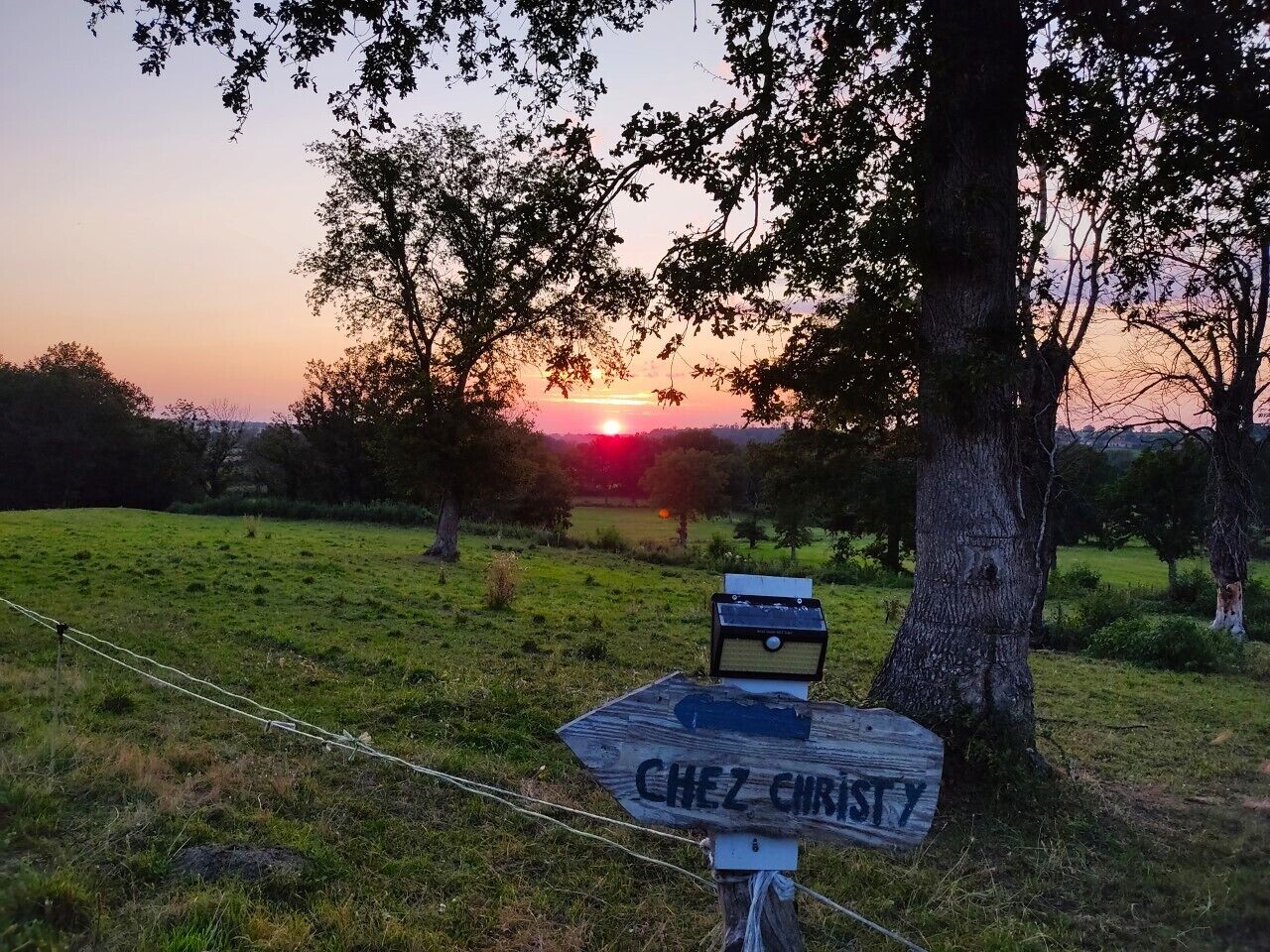 Um letreiro com a inscrição 'CHEZ CHRISTY', ao fundo um pôr do sol com árvores e um campo gramado.