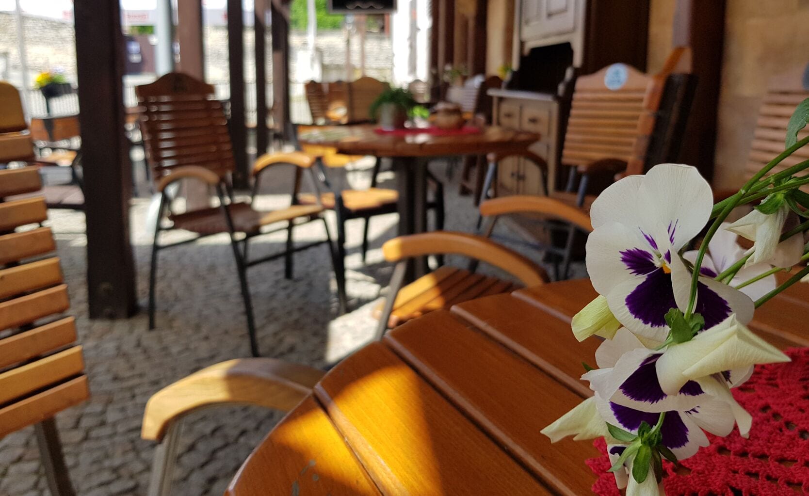 Um café ao ar livre com mesas e cadeiras de madeira, uma pequena mesa com uma planta e um pano vermelho sobre a mesa.