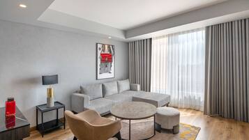 Uma sala de estar moderna com um sofá cinza, uma mesa de centro redonda e uma poltrona bege. Há uma mesinha de apoio com uma luminária e um objeto decorativo vermelho. Uma moldura com um quadro está pendurada na parede, e o quarto possui grandes janelas com cortinas cinzas.