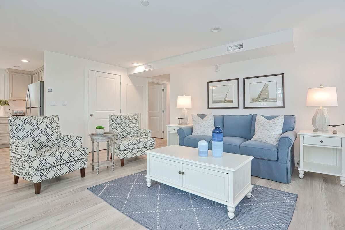 Uma sala de estar moderna com um sofá branco, poltrona estampada e uma mesa de centro. Há duas fotos emolduradas na parede, uma luminária de cada lado do sofá e uma pequena mesa lateral com uma planta.