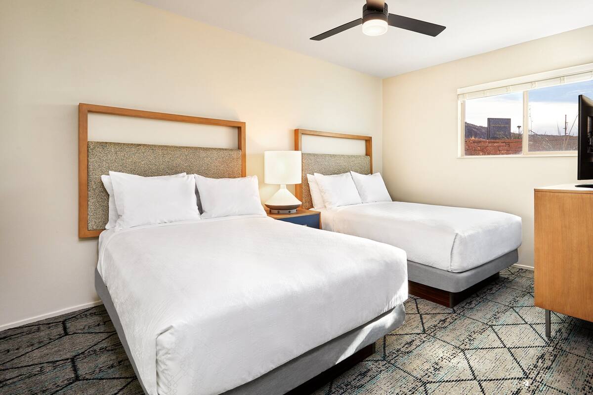 Quarto de hotel com duas camas, um ventilador de teto, mesa de cabeceira com abajur, escrivaninha e janela com vista para uma paisagem desértica.