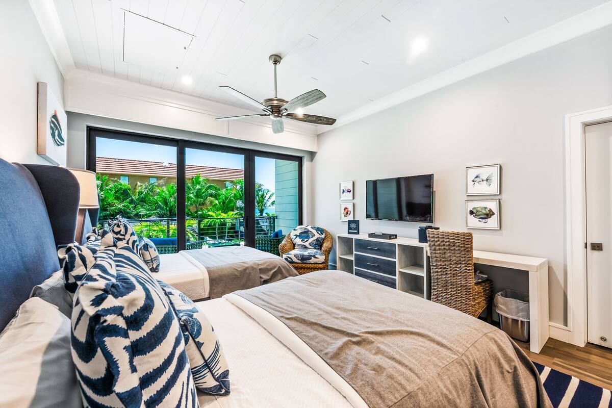 Quarto moderno com duas camas, ventilador de teto, televisão e uma grande janela com vista para o verde.