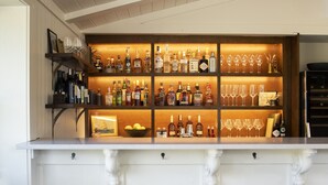 Uma área de bar com balcão de madeira, dois bancos e prateleiras cheias de garrafas e copos.