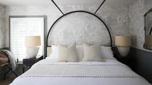 Quarto com cama, duas luminárias de cabeceira, mesa de cabeceira, cadeira e janela com persianas.