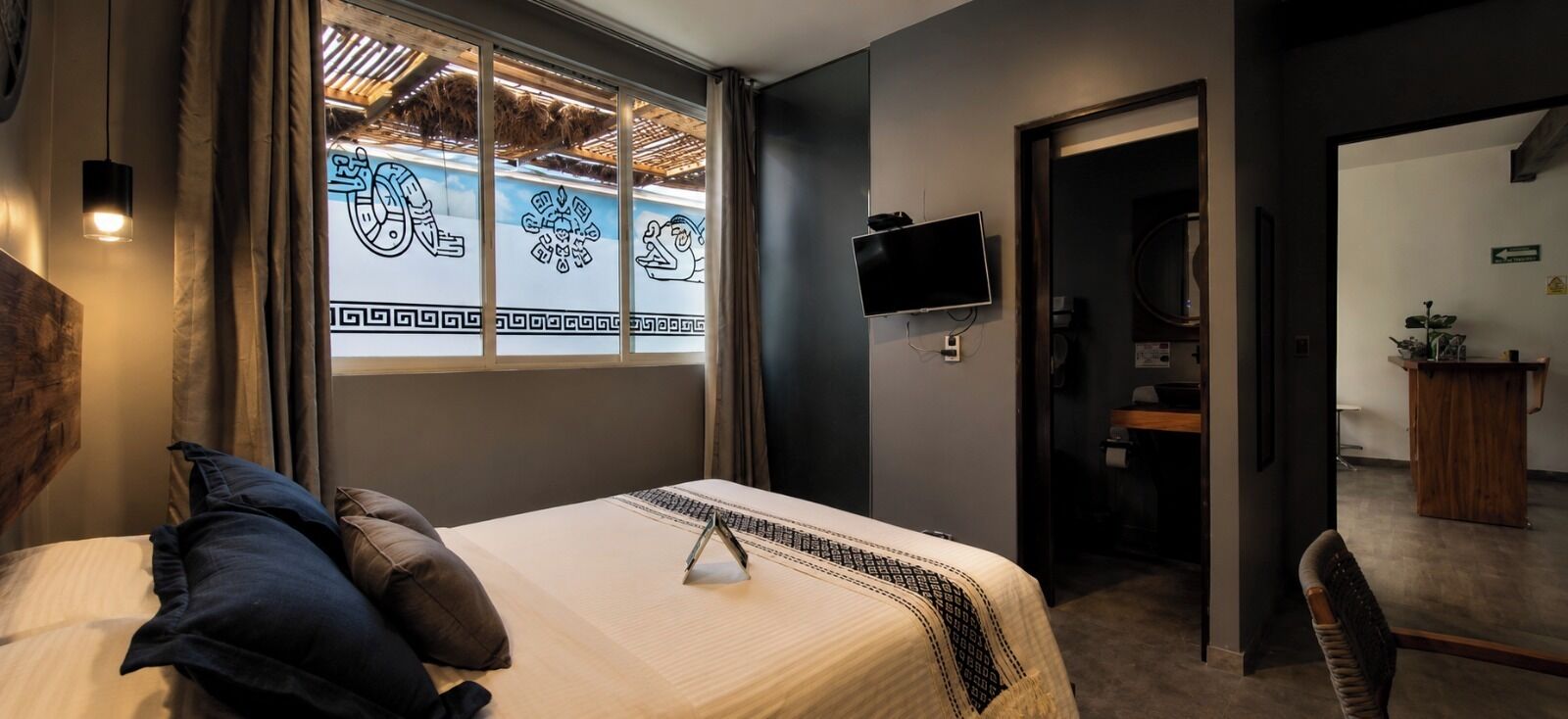 Quarto de hotel com cama, televisão na parede e janela com desenhos decorativos.