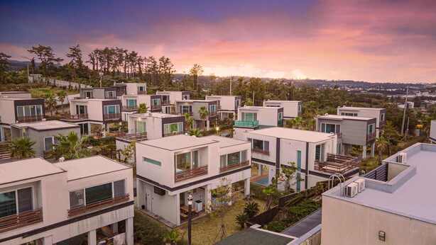 Um conjunto de edifícios residenciais modernos, com várias andares, varandas e painéis solares, cercado por vegetação e uma vista panorâmica do pôr do sol.