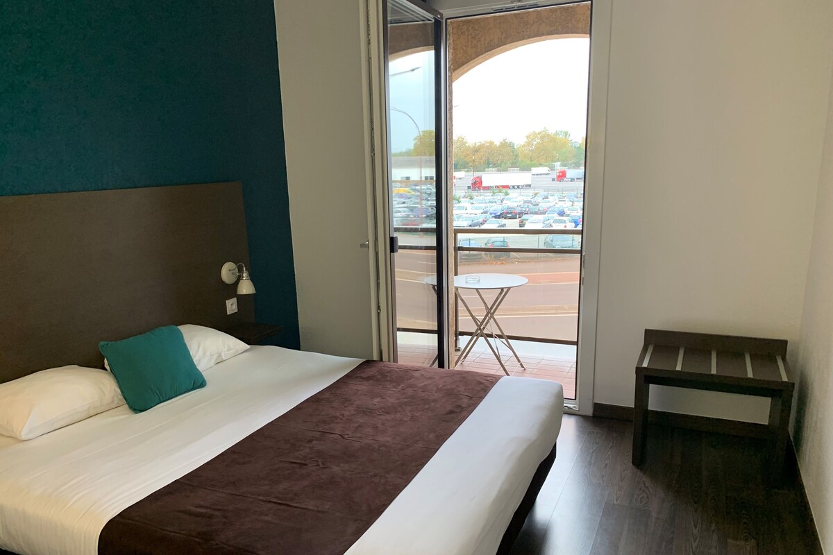 Quarto de hotel com uma cama, uma mesinha e vista para um estacionamento através de uma porta de vidro.