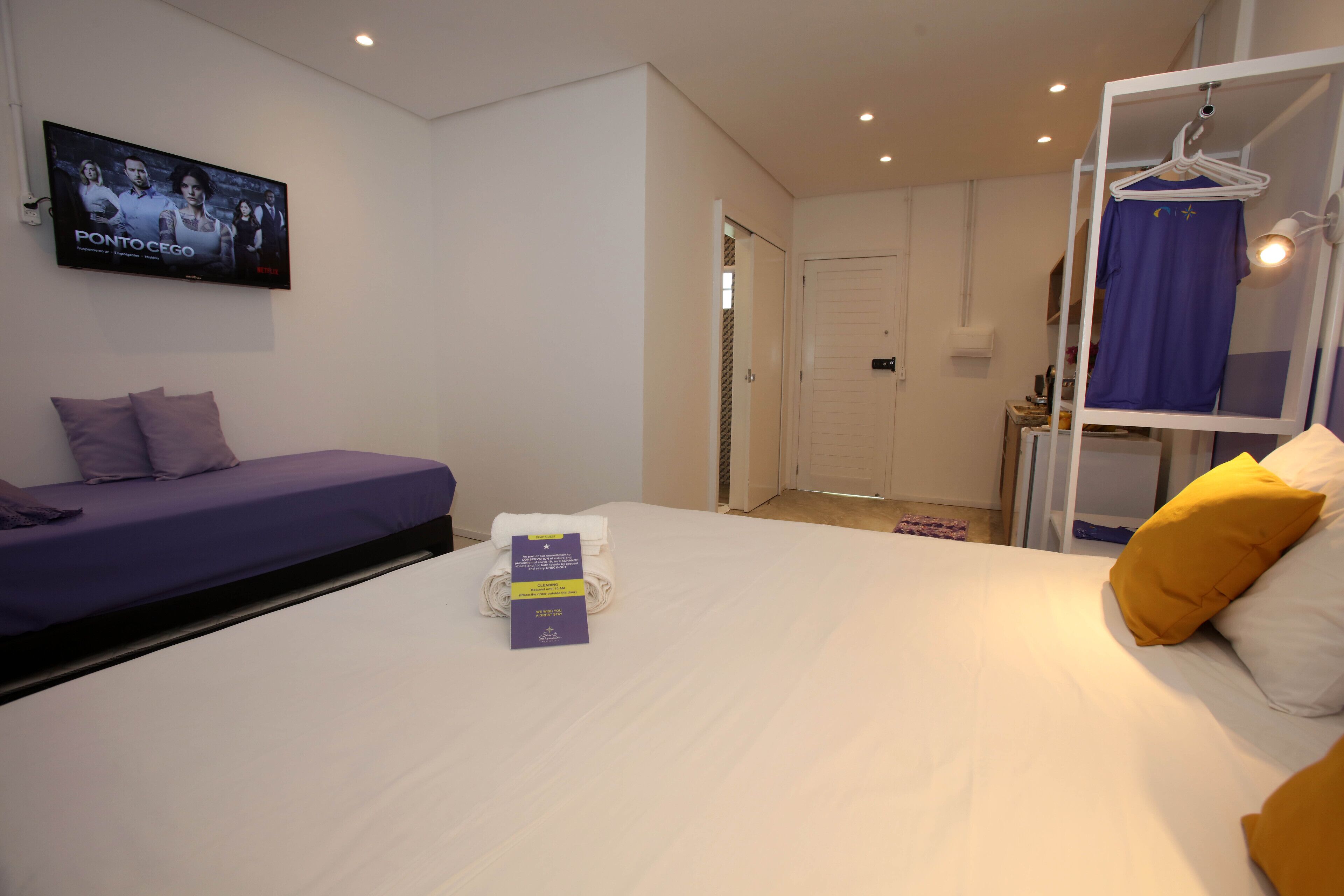 Quarto de hotel com uma cama, televisão fixada na parede e um cabideiro com roupas.