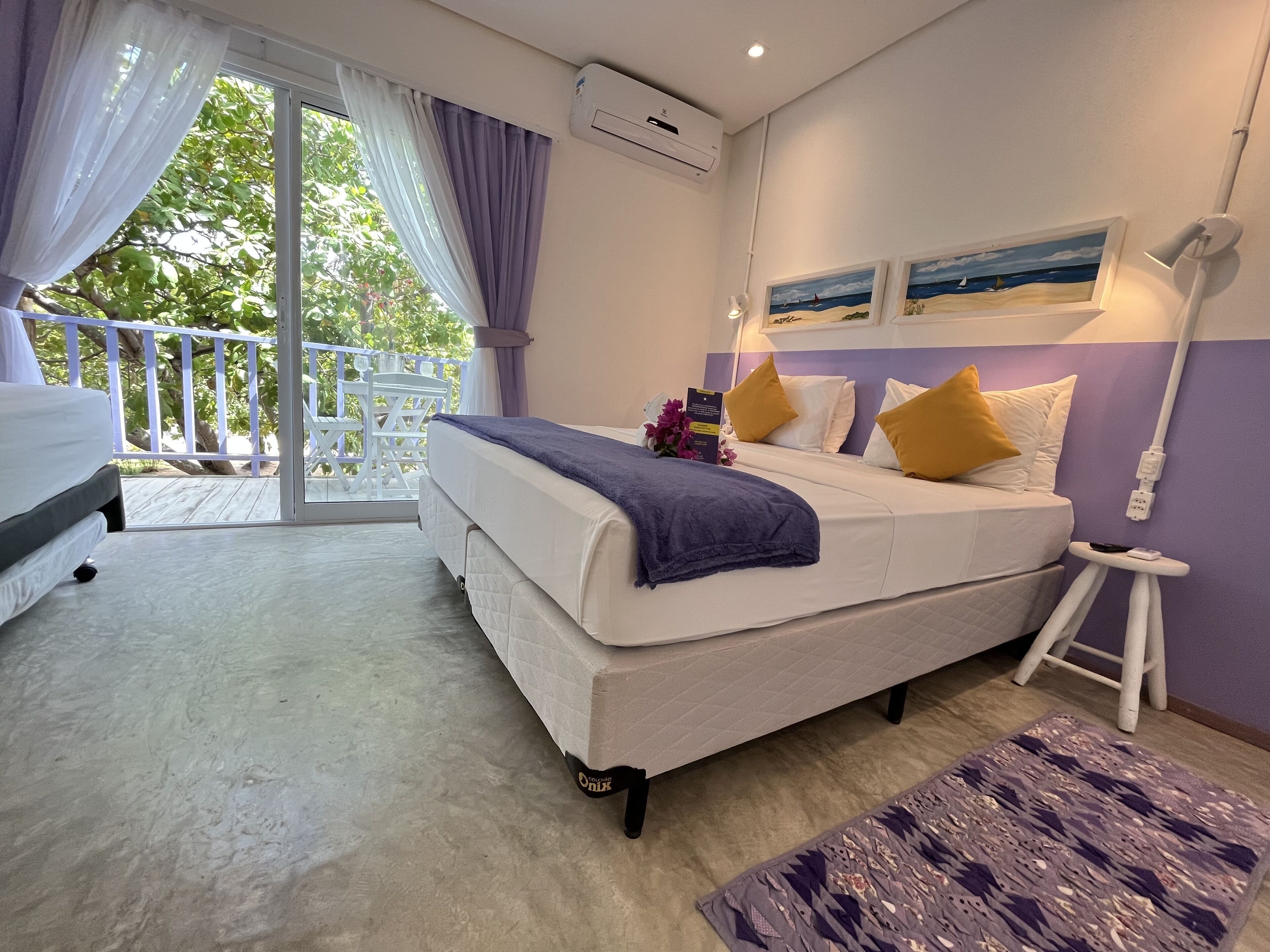 Quarto com cama, uma mesinha, criado-mudo, janela com cortinas, ar condicionado e um quadro na parede.