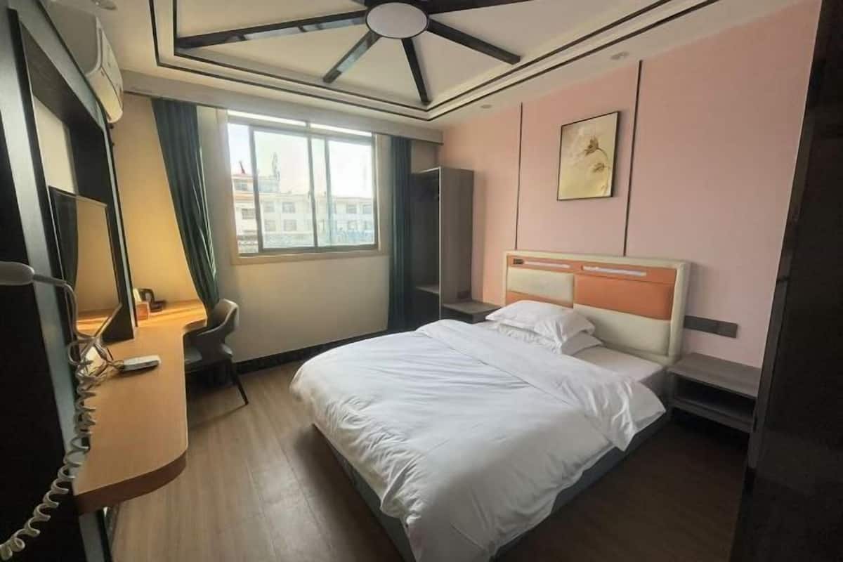 Quarto de hotel com cama, escrivaninha com telefone, janela com cortinas e ventoinha no teto.