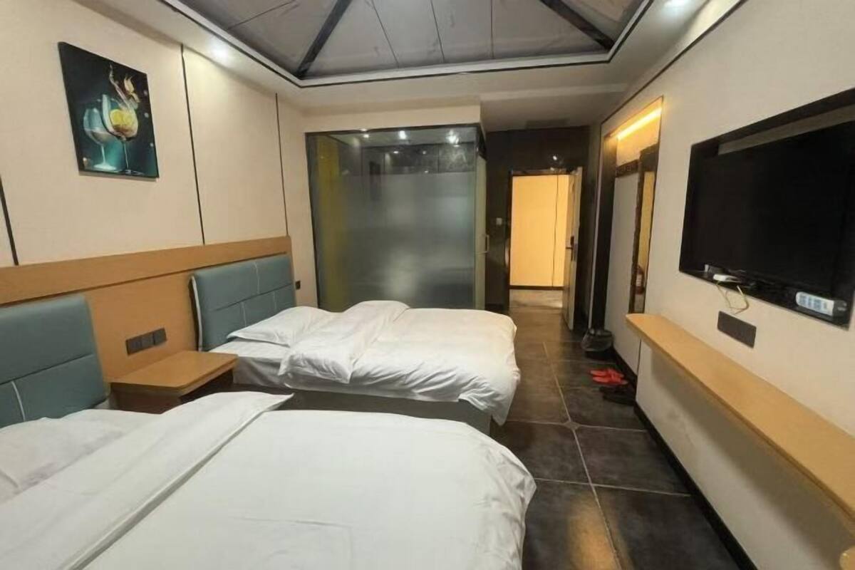 Quarto de hotel com duas camas, uma televisão, um quadro e uma porta de vidro que dá para um corredor.