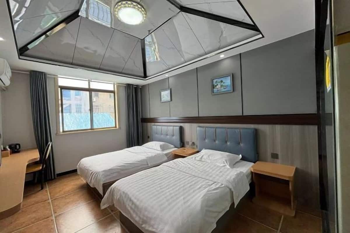 Quarto de hotel com duas camas, uma escrivaninha, uma cadeira, uma janela com cortinas, uma luminária no teto e um quadro na parede.