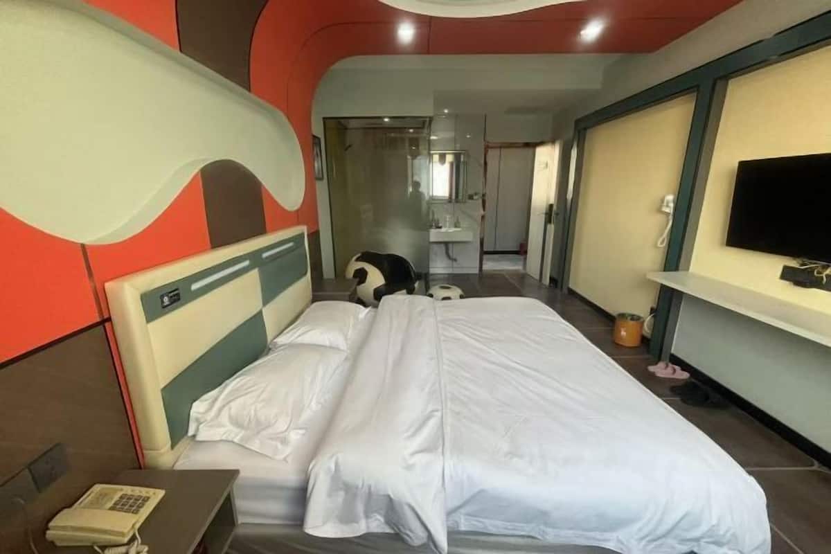 Quarto de hotel com cama, mesa de cabeceira com telefone, televisão na parede e banheiro visível por uma porta aberta.