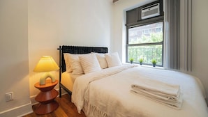 Cama bem arrumada com radiador, abajur e janela com vista para prédios e vegetação.