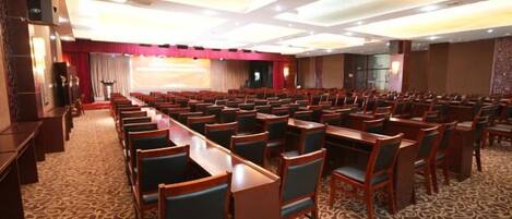 Uma sala de conferência com fileiras de mesas e cadeiras, um palco com cortinas e piso coberto por carpete.