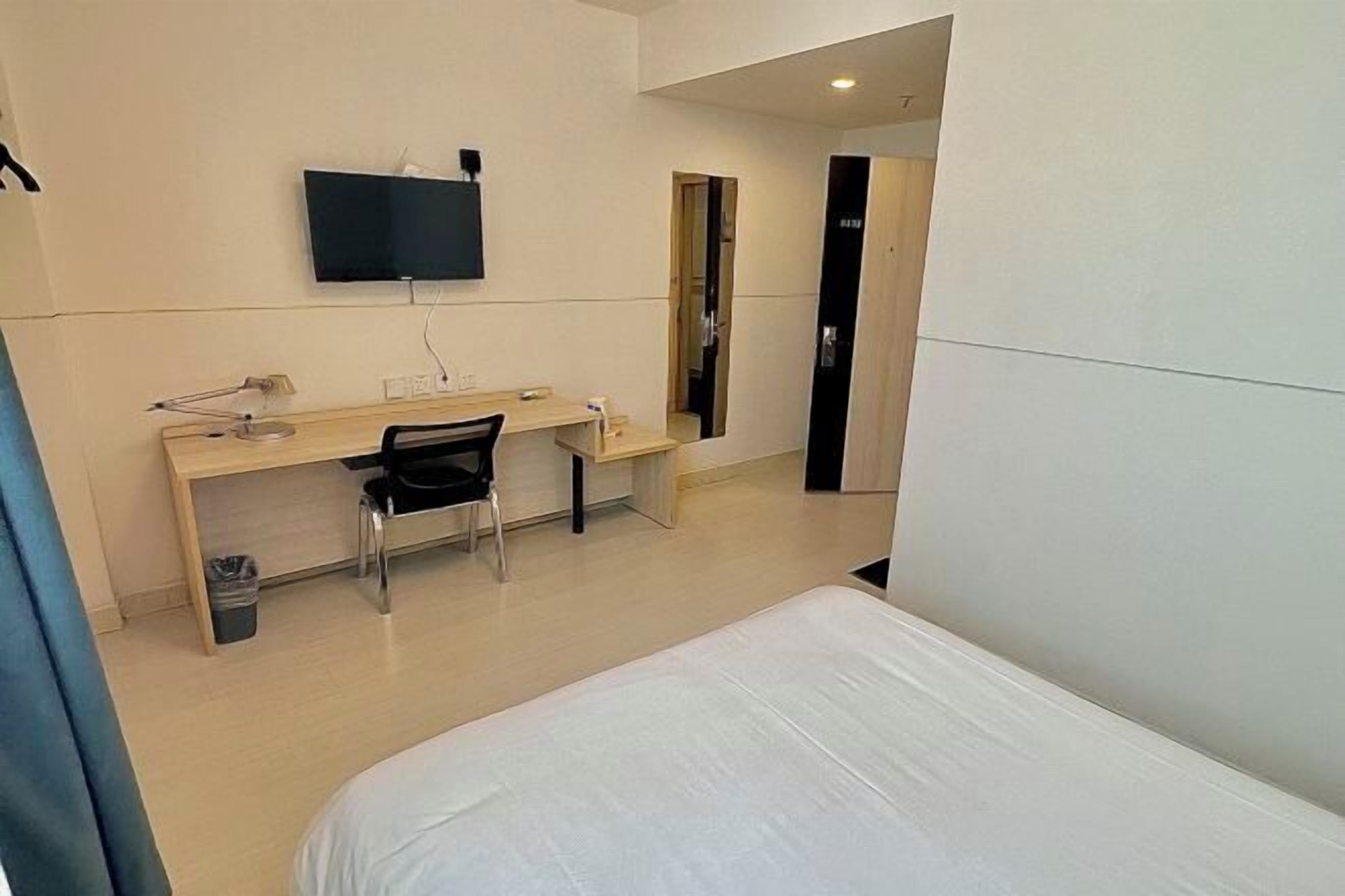 Quarto de hotel com uma cama, uma escrivaninha, uma cadeira, uma televisão fixada na parede e uma porta que dá para outro quarto.