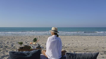 Uma pessoa sentada numa cadeira de praia, com uma mesa de comida e bebidas à frente, com vista para o oceano.