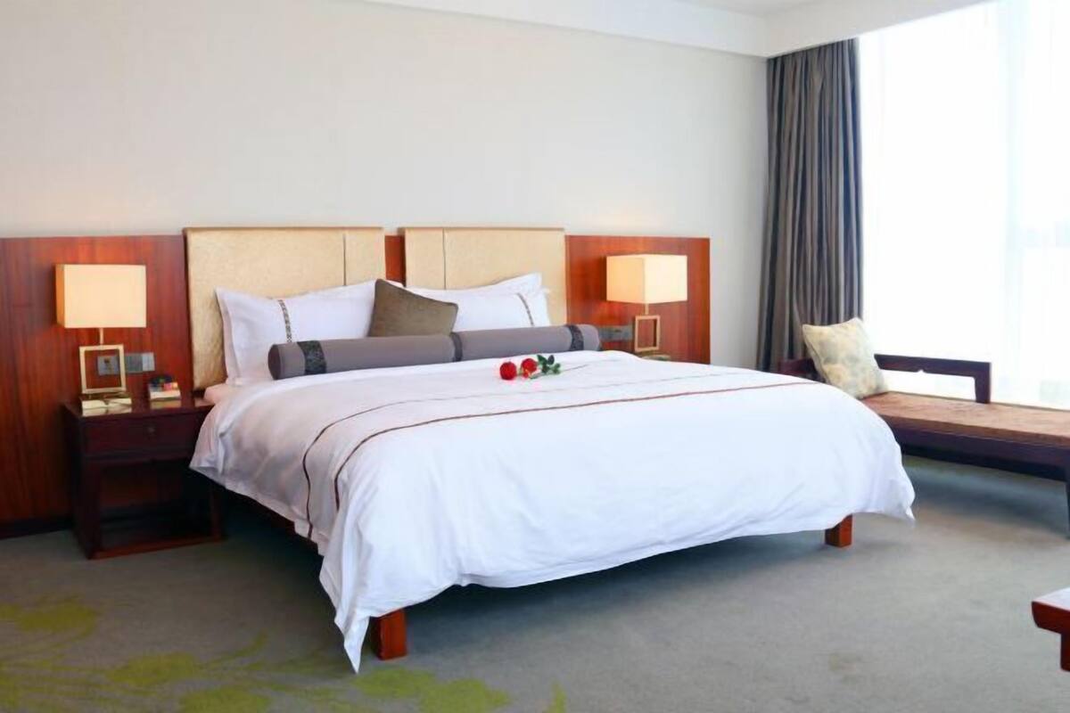 Quarto de hotel com uma cama grande, duas mesas de cabeceira, um banco e uma janela com cortinas.