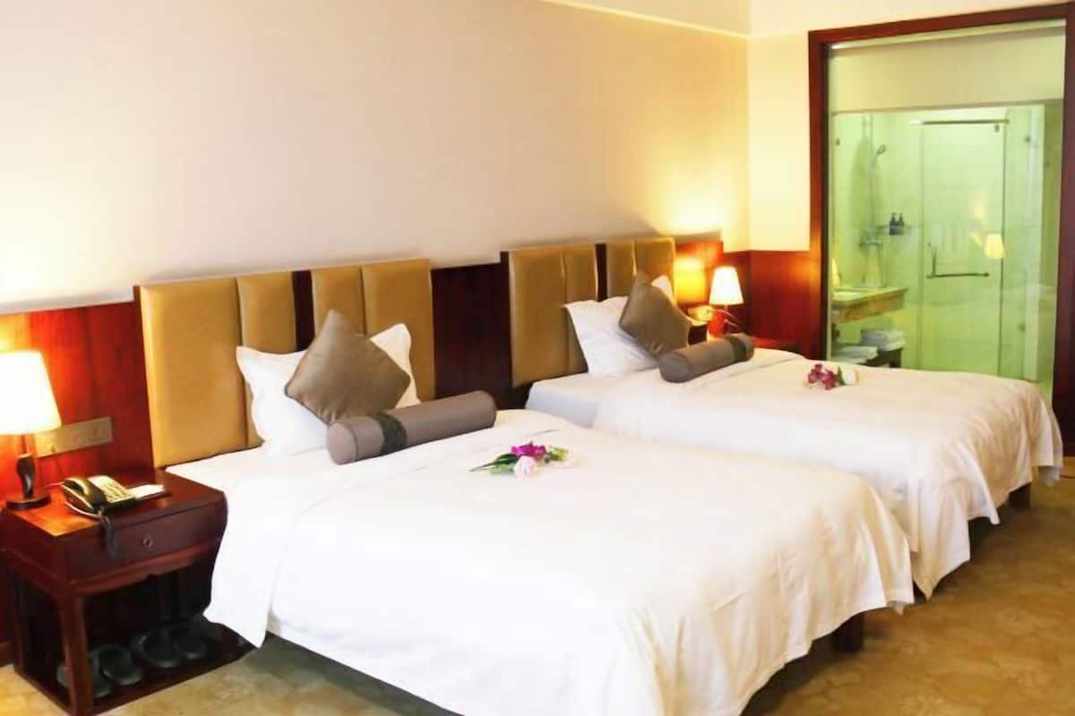 Quarto de hotel com duas camas, mesa de cabeceira com telefone, e banheiro visível por uma porta de vidro.