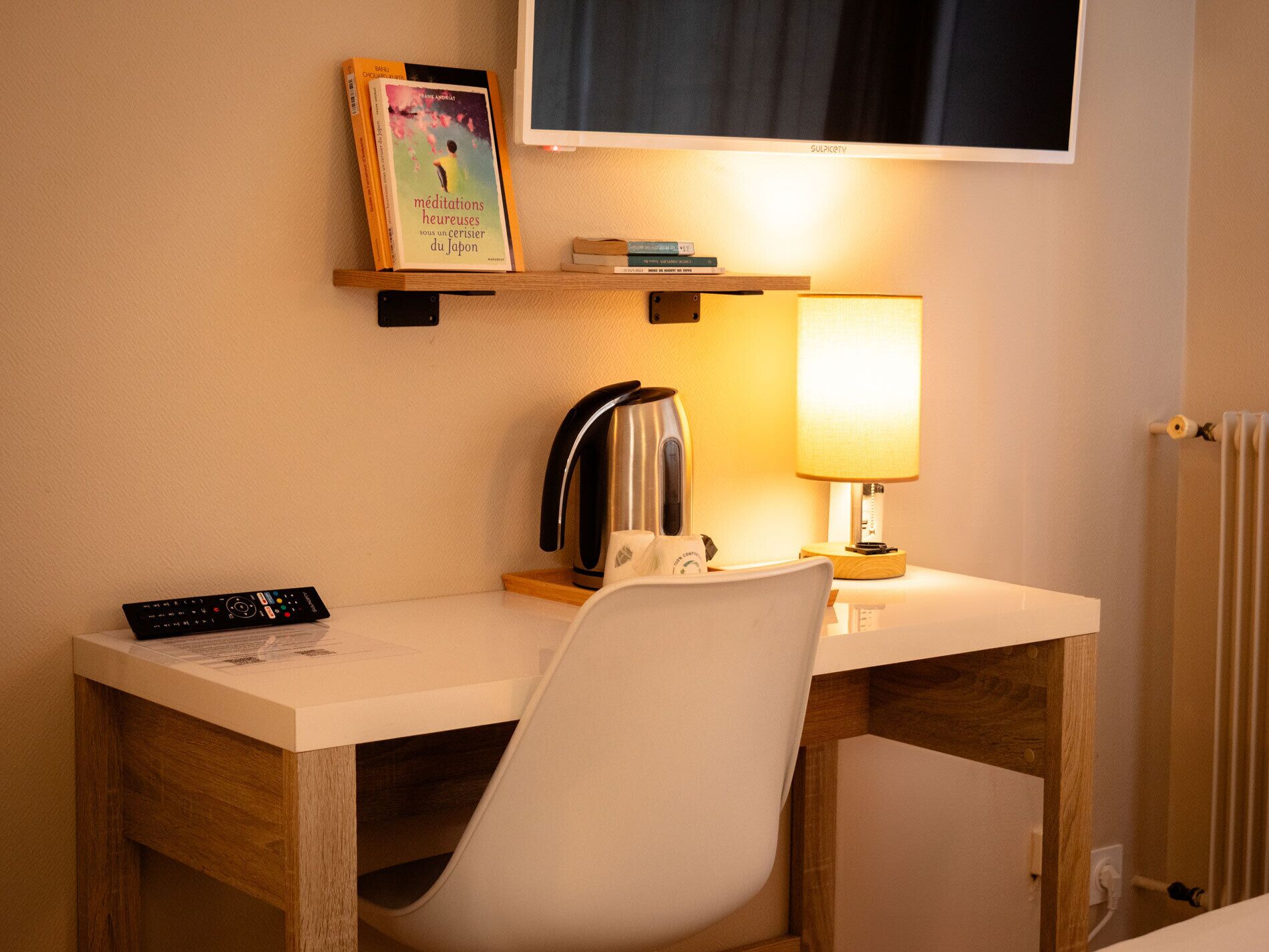 Quarto de hotel com escrivaninha, cadeira, chaleira, controle remoto, luminária e televisão.