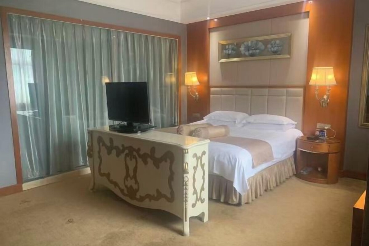 Quarto de hotel com cama, televisão em suporte, mesa de cabeceira com abajur e uma janela grande com cortinas.