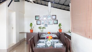 Uma sala de jantar com uma mesa comprida posta para quatro pessoas, rodeada por cadeiras. Há plantas em vasos em ambos os lados da mesa, e uma fileira de quadros na parede atrás da mesa.