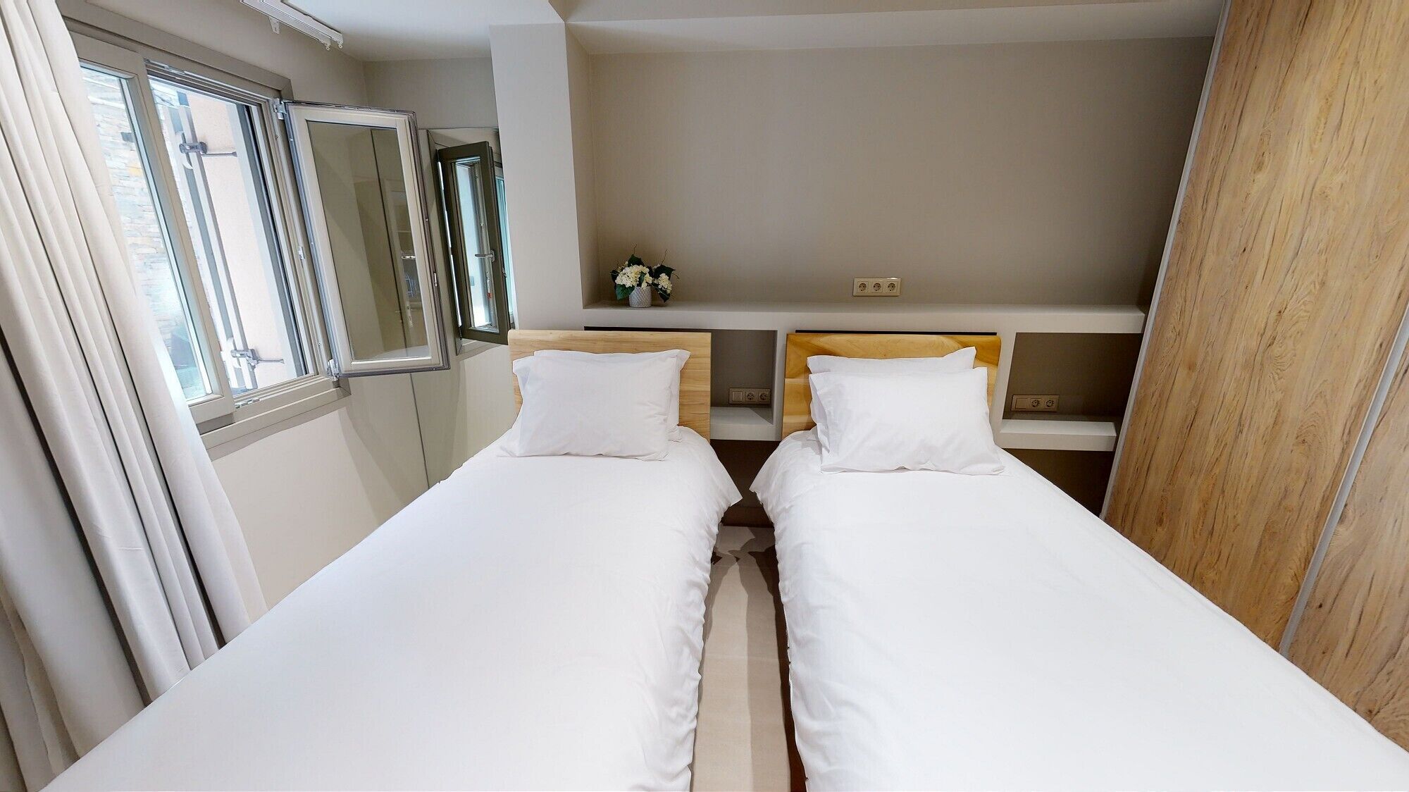 Quarto de hotel com duas camas de solteiro, uma mesinha com um vaso de flores e uma janela com cortinas.