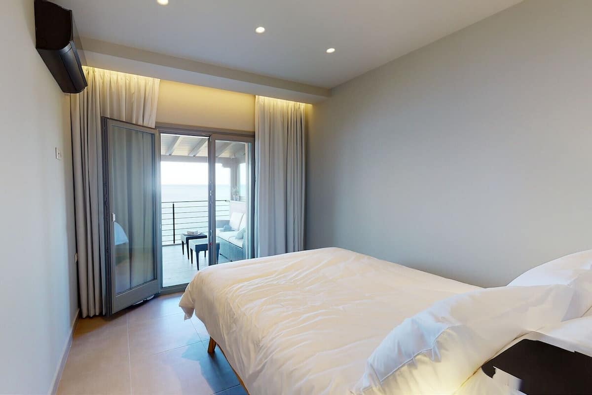 Quarto de hotel com uma cama, televisão fixada na parede e porta de vidro deslizante para uma varanda com mesa e cadeiras.