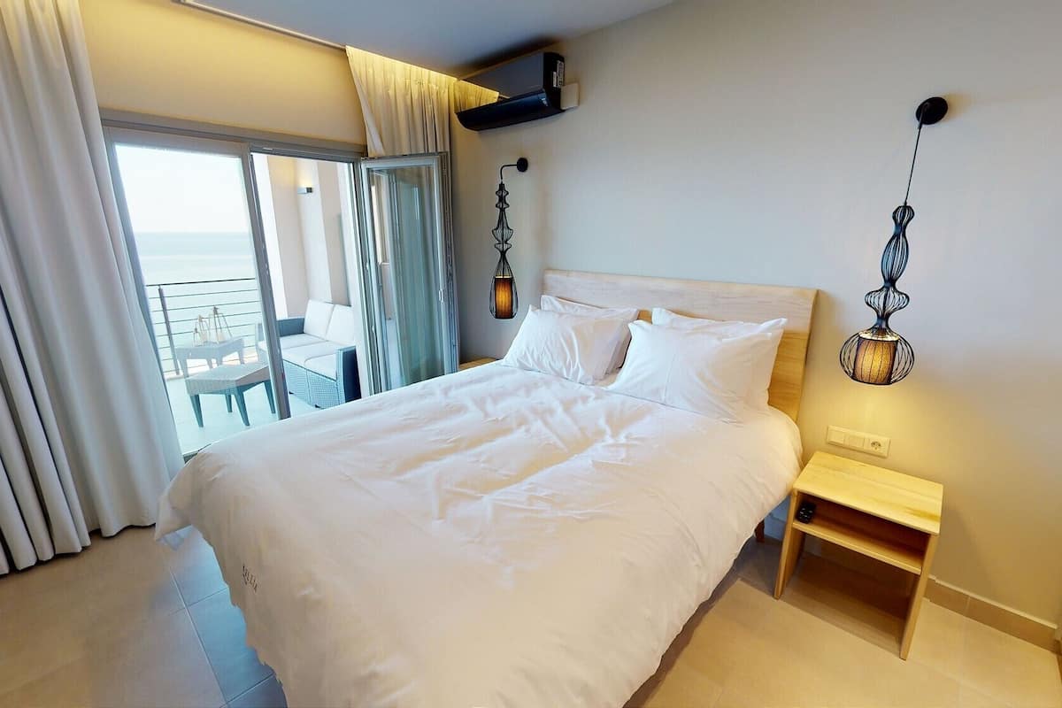 Quarto de hotel com cama grande, mesa de cabeceira, luminária na parede e vista para a varanda.