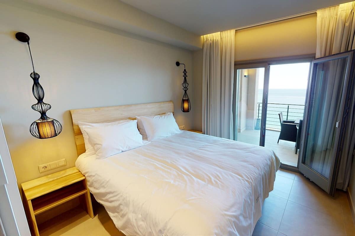 Quarto de hotel com uma cama grande, mesas de cabeceira, abajur e vista para o mar através de uma porta de vidro deslizante.