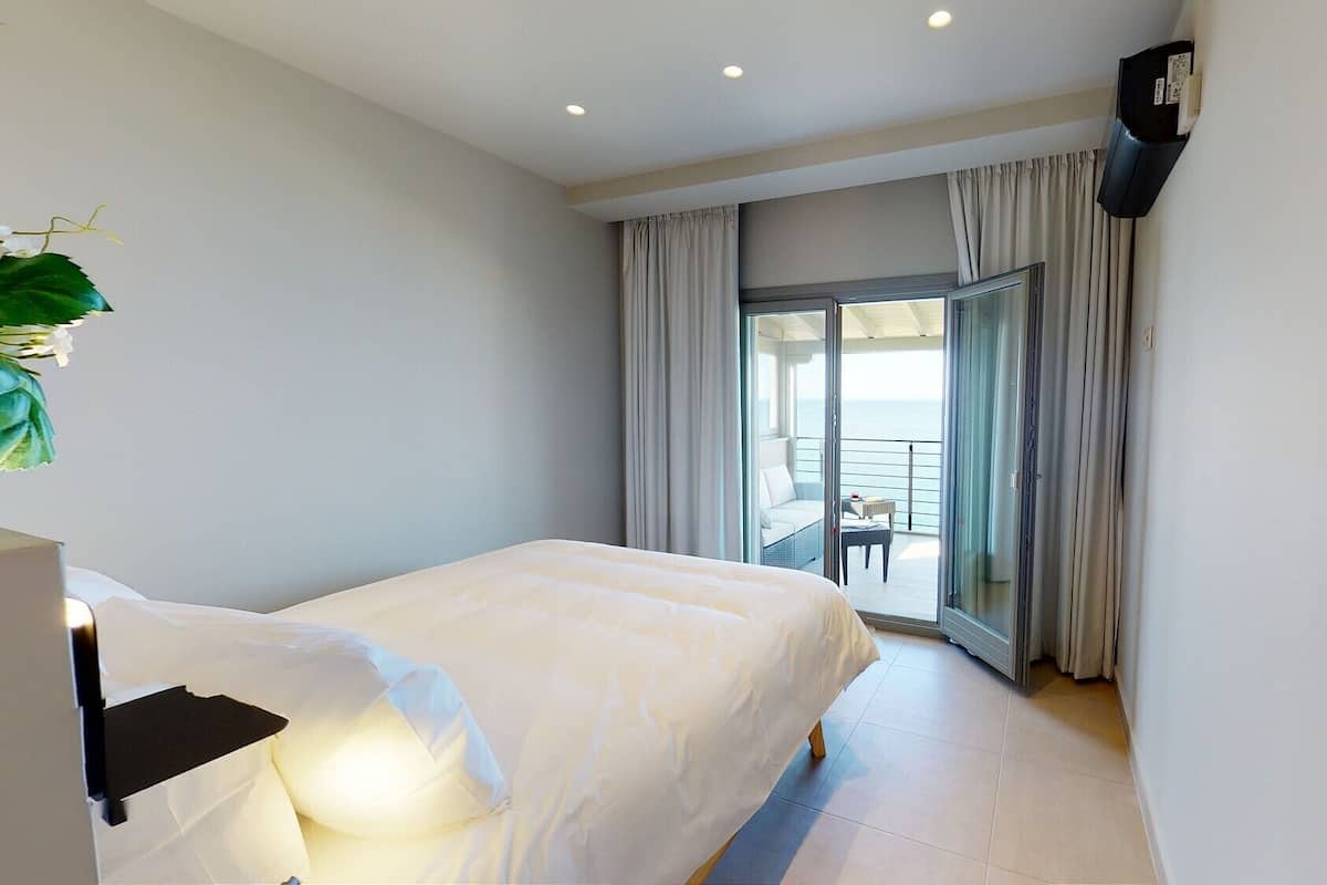 Quarto de hotel com cama, uma mesinha, uma cadeira e vista para o mar através de uma porta de vidro.