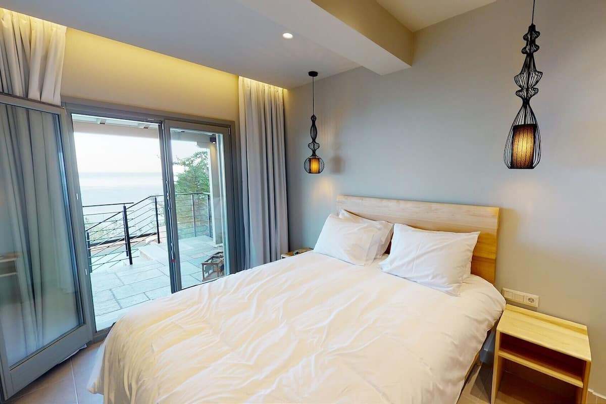 Quarto de hotel com uma cama grande, uma mesa de cabeceira, uma luminária na parede e uma porta de vidro deslizante que dá para uma varanda com vista para o mar.
