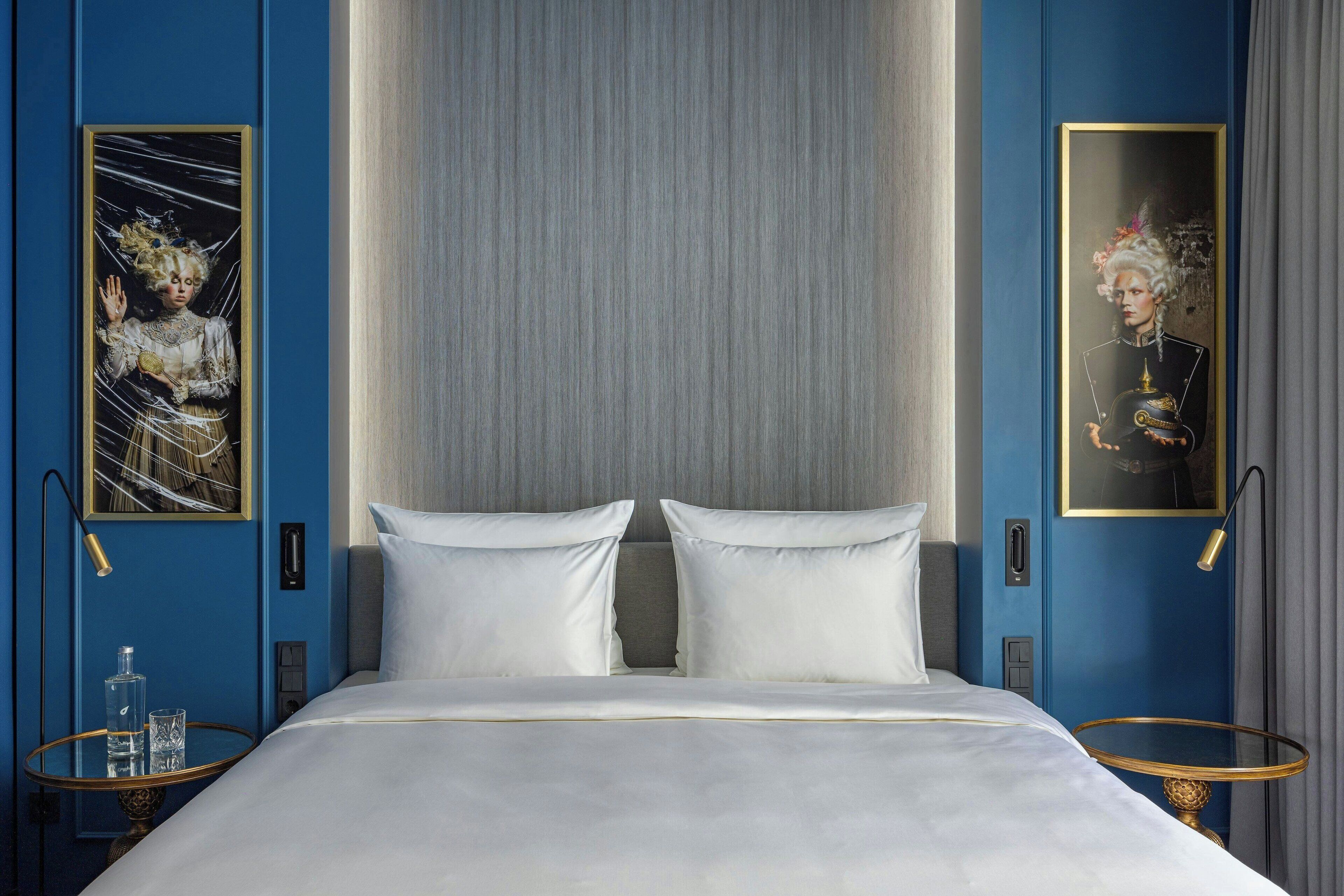 Quarto de hotel com uma cama grande, duas almofadas brancas, uma mesa de cabeceira com uma garrafa e um copo, e duas fotos emolduradas nas paredes.