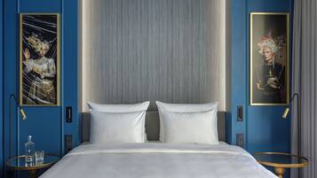 Quarto de hotel com uma cama grande, duas almofadas brancas, uma mesa de cabeceira com uma garrafa e um copo, e duas fotos emolduradas nas paredes.