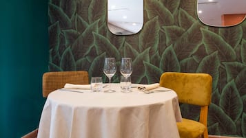 Uma mesa de jantar com toalha branca, dois copos de vinho e um copo pequeno de água. Há duas cadeiras, uma com padrão xadrez e outra de cor lisa. A parede atrás da mesa tem um padrão de folhas.